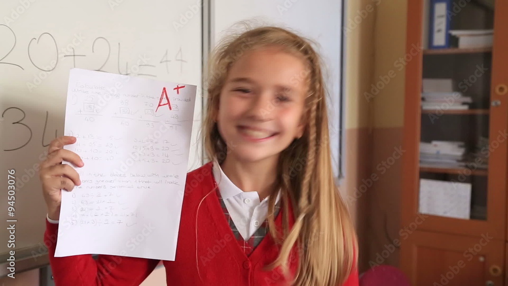 Happy cute little girl showing exam paper with a plus in the classroom ...