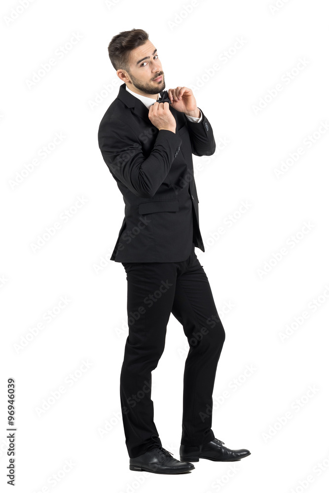Side view of cool elegant young male model adjusting bow tie looking at ...
