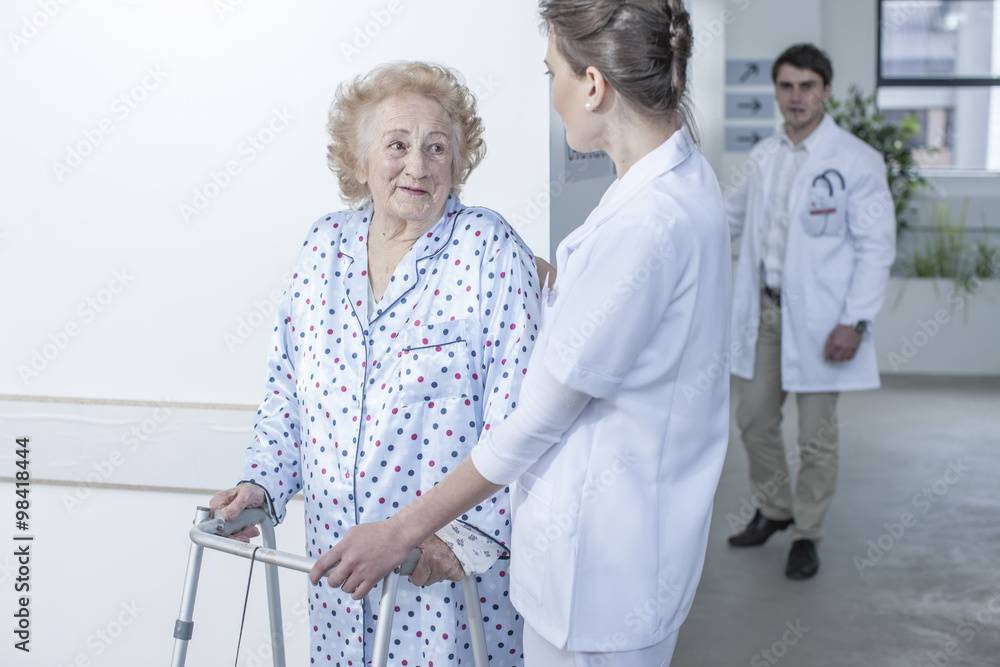 Nurse leading elderly patient with walking frame on hospital floor ...
