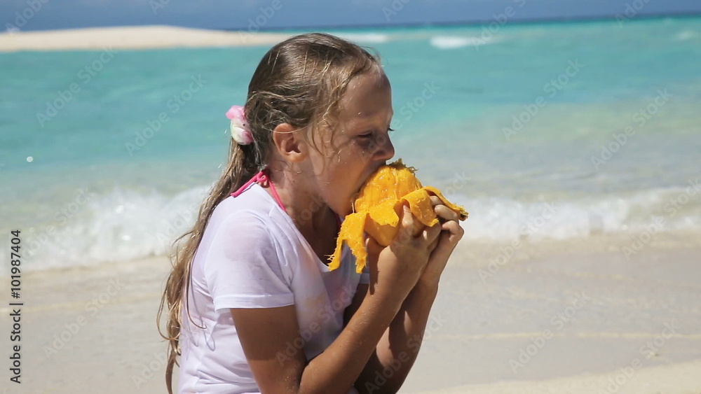 Young girl sitting on a tropical beach, cleans mango peel, eat mango ...