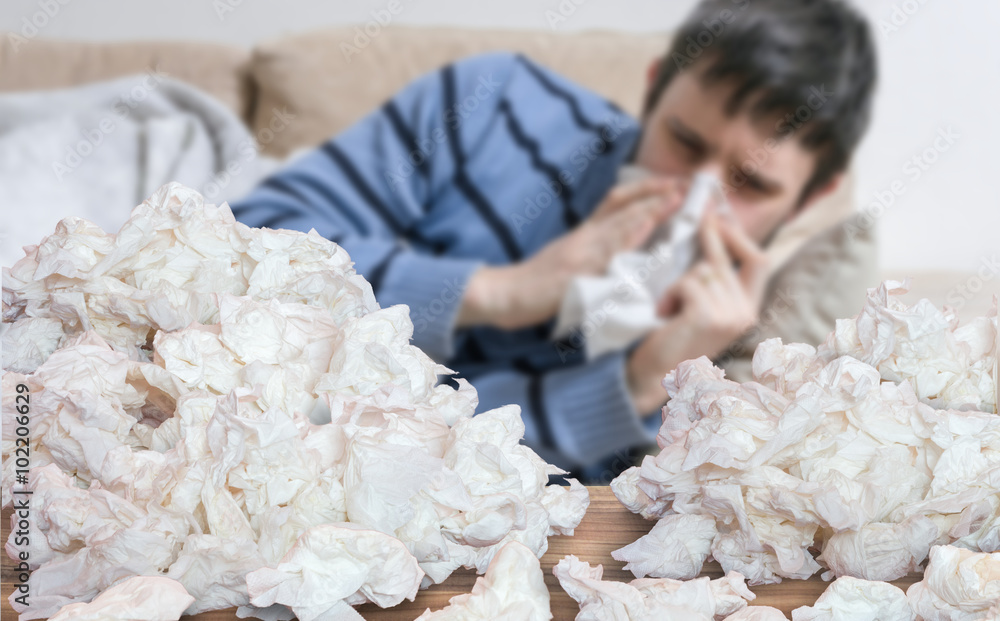 Funny sick man who has flu or cold is blowing his nose. Pile of tissues ...