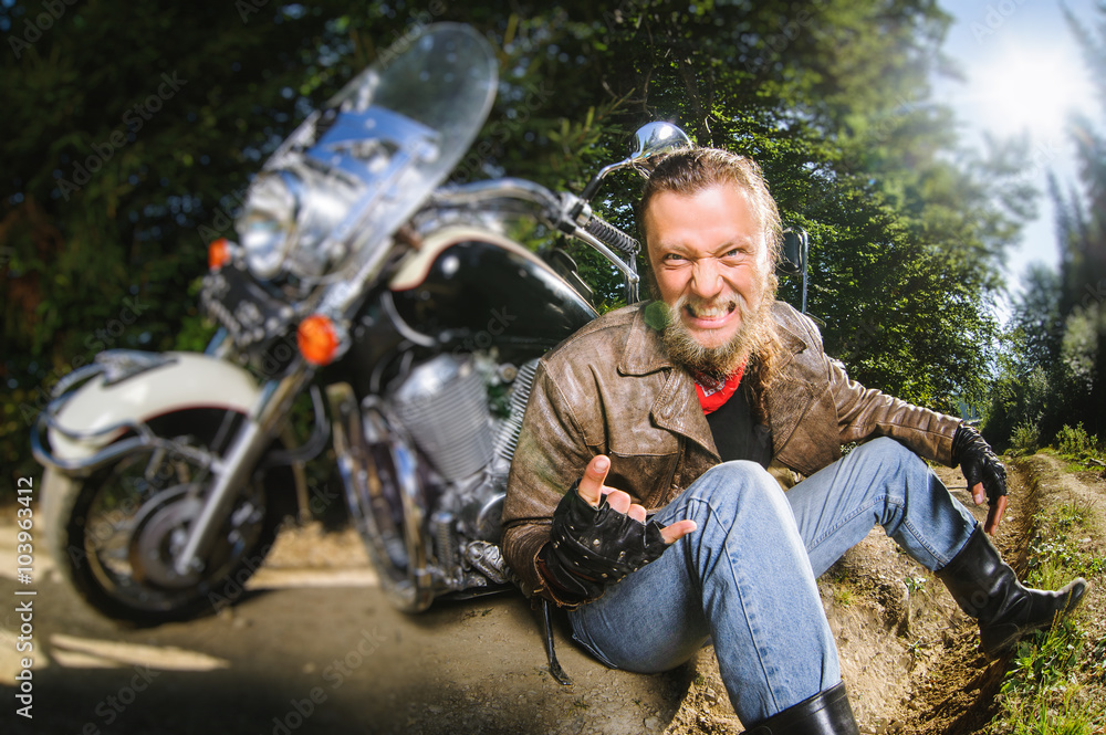 Unshaven male biker in leather jacket and blue jeans sitting on road ...