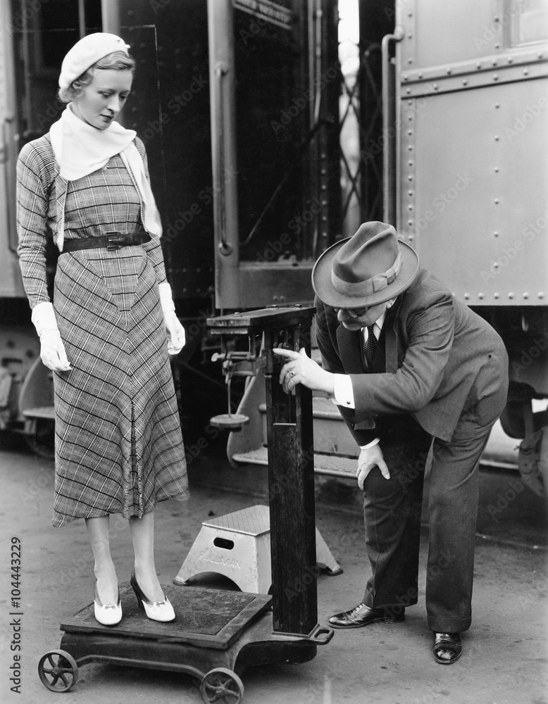 Profile of a man measuring weight of a woman standing on a weighing ...