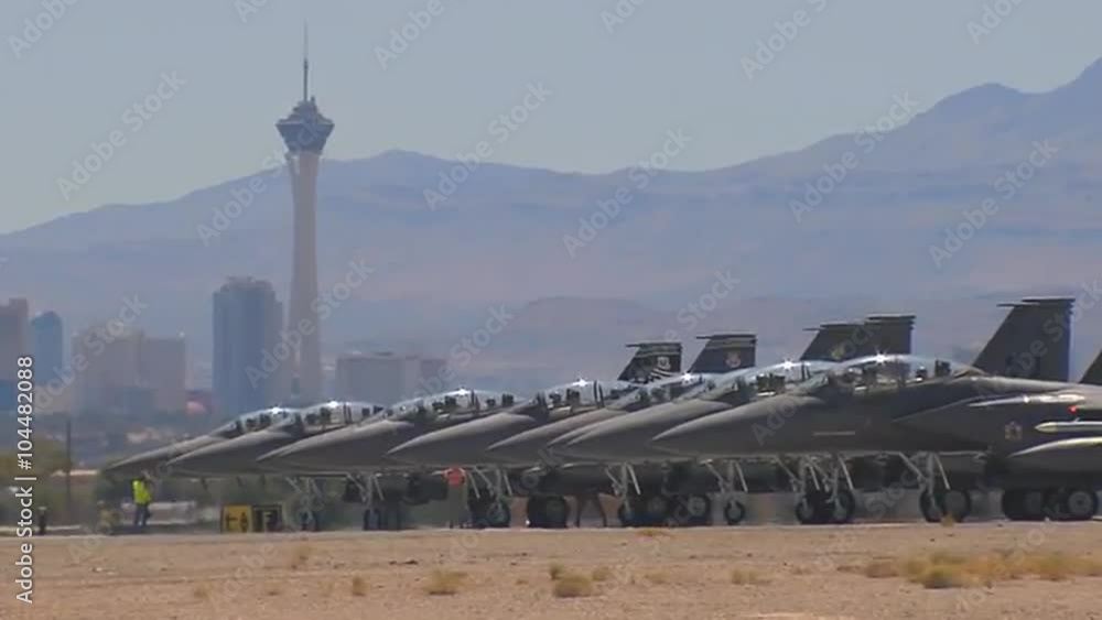 Numerous F-15 and F-16 fighter jets line up and taxi for takeoff in a ...