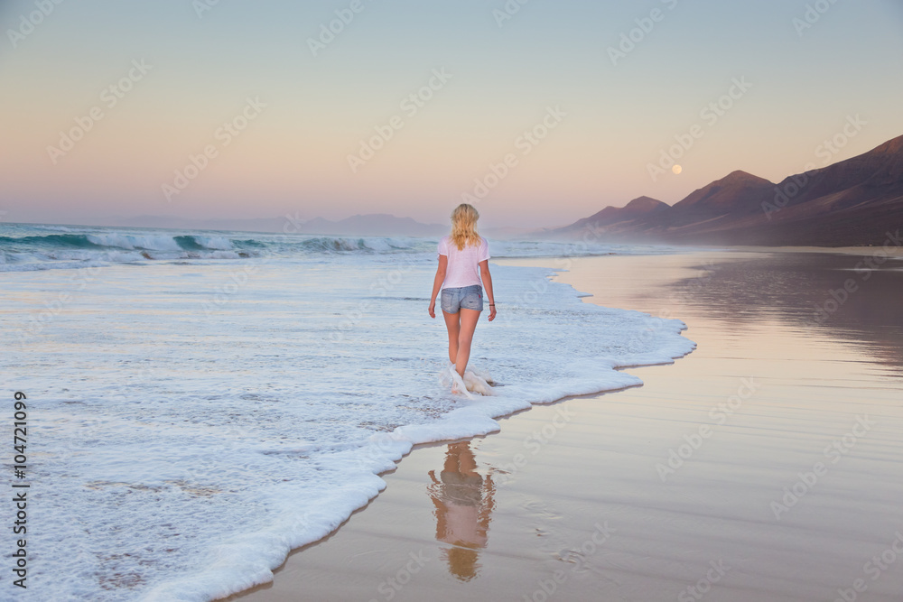 Woman walking on sandy beach in sunset. Waves sweeping away her traces ...