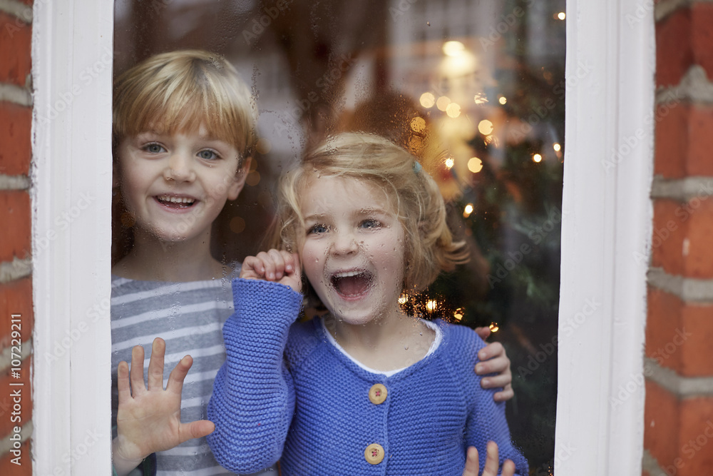 Two children, a boy and girl, looking out of a window at home with ...