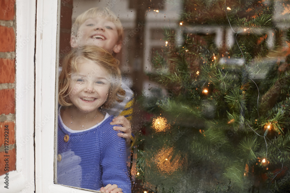 Two children, a boy and girl, looking out of a window at home beside a ...