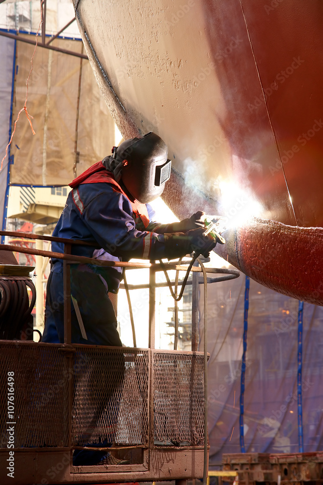 Shipyard welder welding the hull of the ship at the dock with lift ...