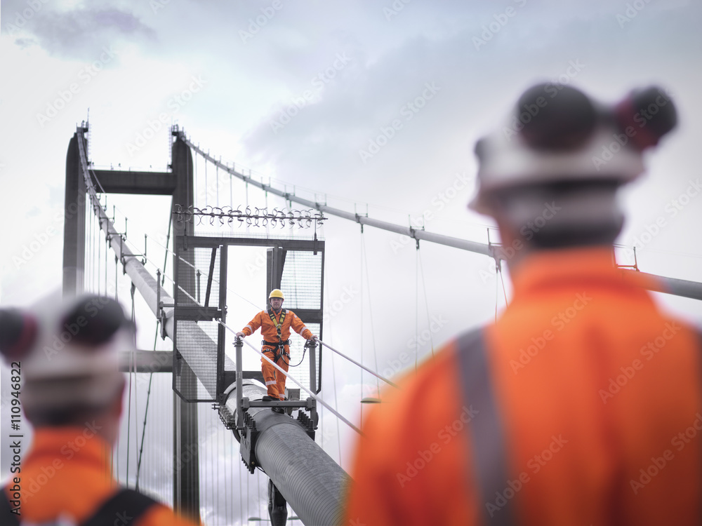 Bridge workers watching coworker on cable of suspension bridge. The ...
