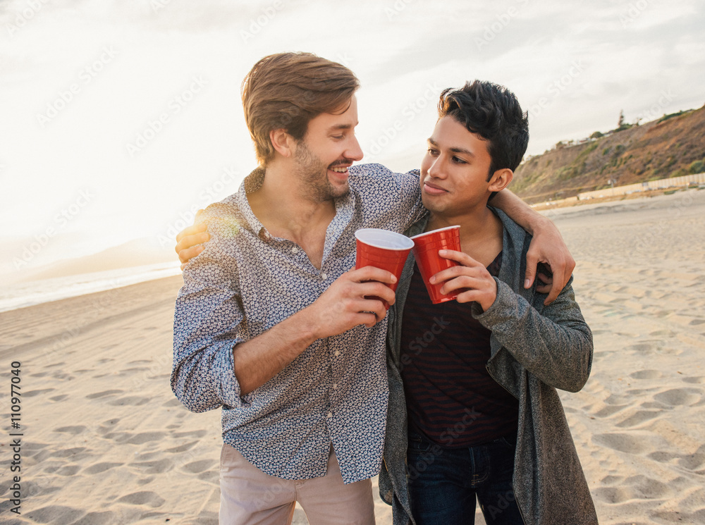 Two male friends having drinks together on beach, arms around each ...