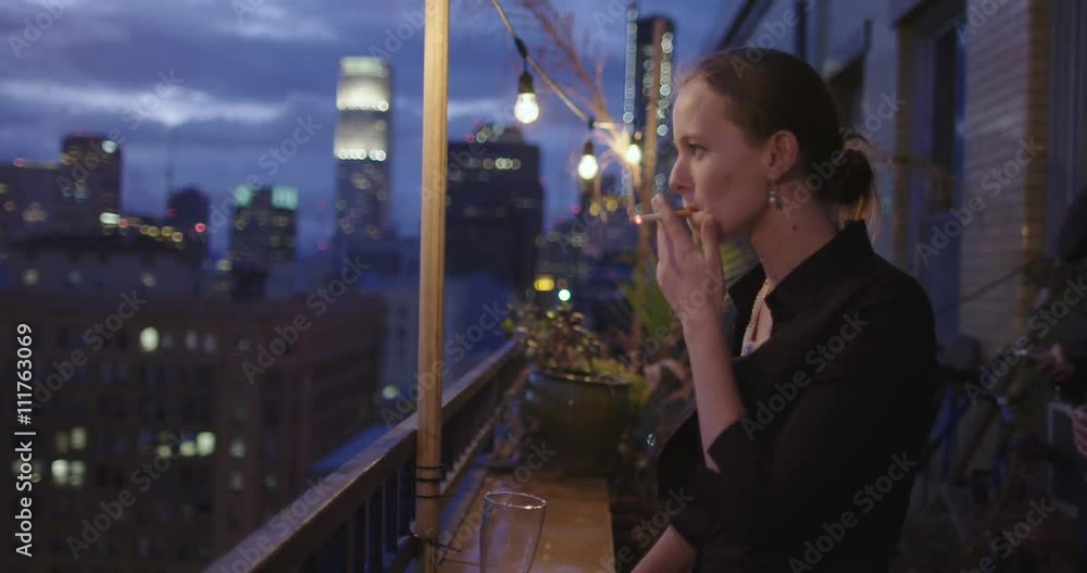 Attractive young woman smokes a cigarette in front of the night skyline ...