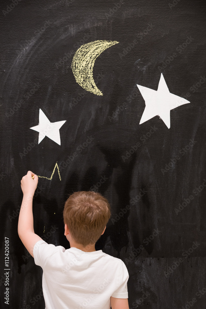 Back view on cute kid boy drawing stars with chalk on a black board ...