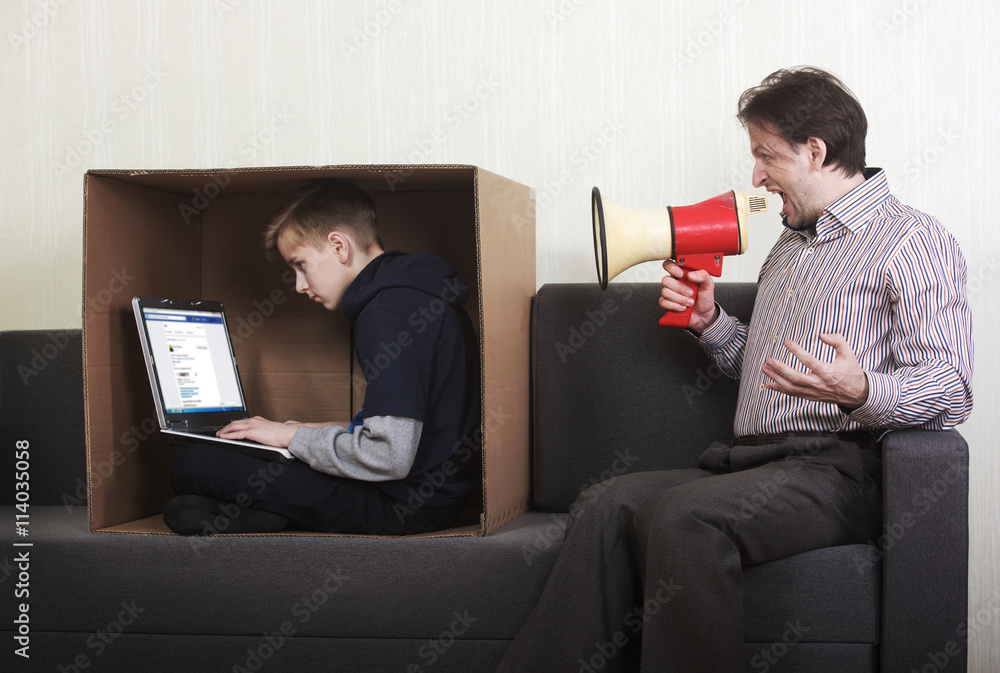 Tween son sitting in a cardboard box with a laptop while his father ...