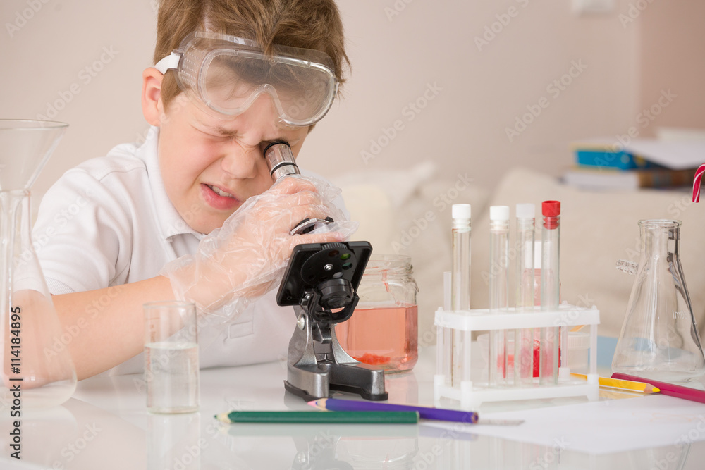 Cute elementary schoolboy looking into microscope at his desk at home ...