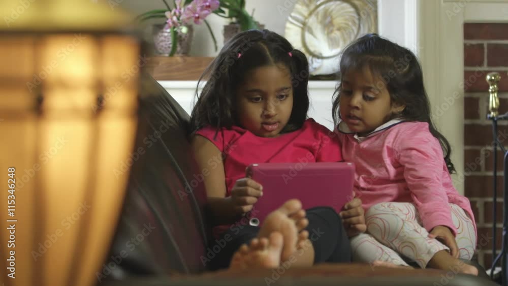 Scene rack focuses to cute little Indian sisters on a couch captivated ...