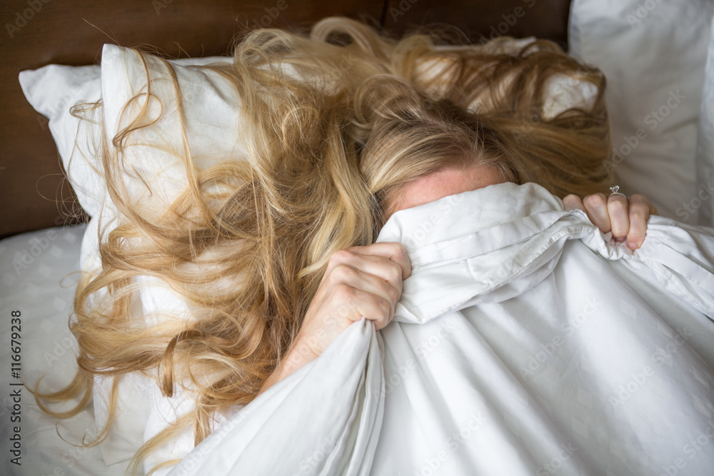 Scared young woman lying in bed covered with a blanket and screaming ...