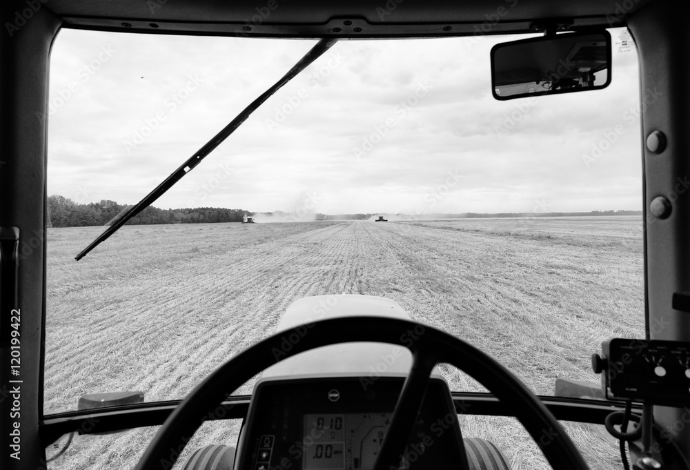 A tractor window framing a field of wheat being harvested by two ...