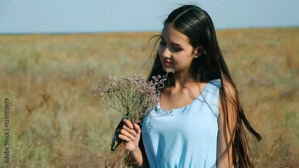 Young beautiful girl in a blue dress enjoys flowers steppe. A young ...