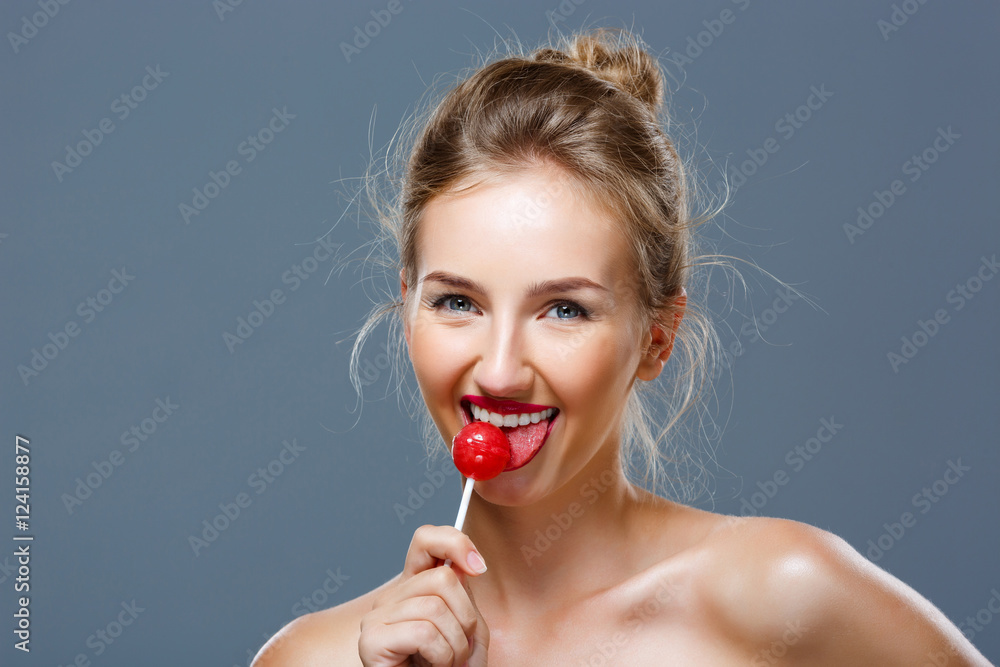 Young Beautiful Blonde Girl Eating Chupa Chups Over Grey Background