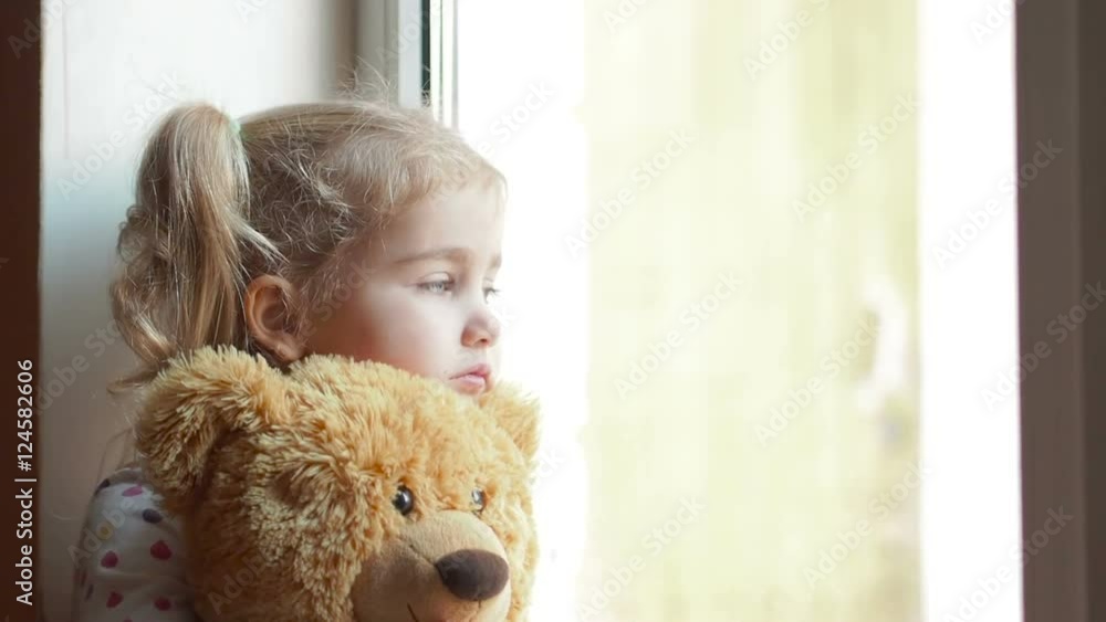 Little girl stares sadly out of a window with a teddy bear in her arms ...