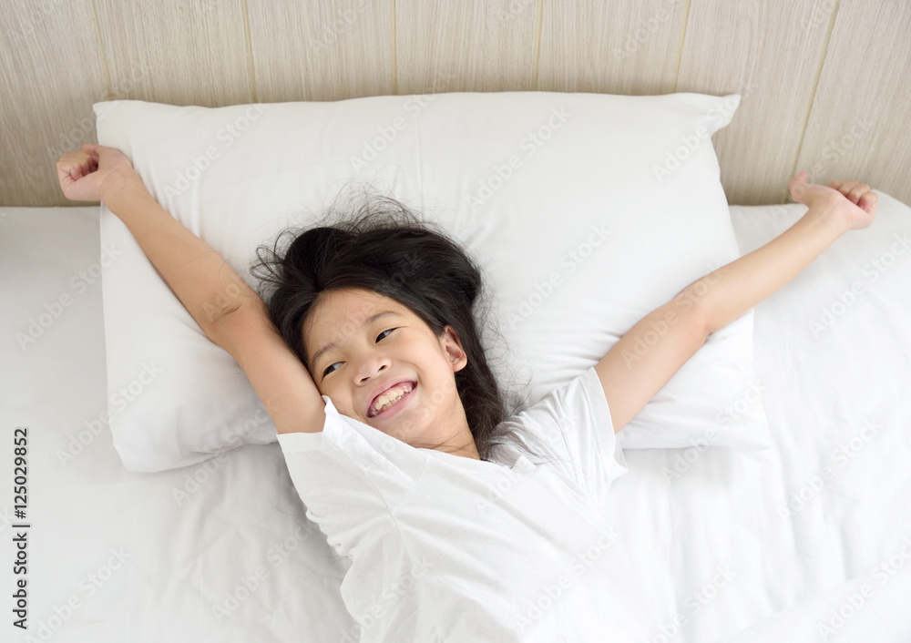Portrait of happy young Asian girl lying in bed after waking up and stretching arms on the bed ...