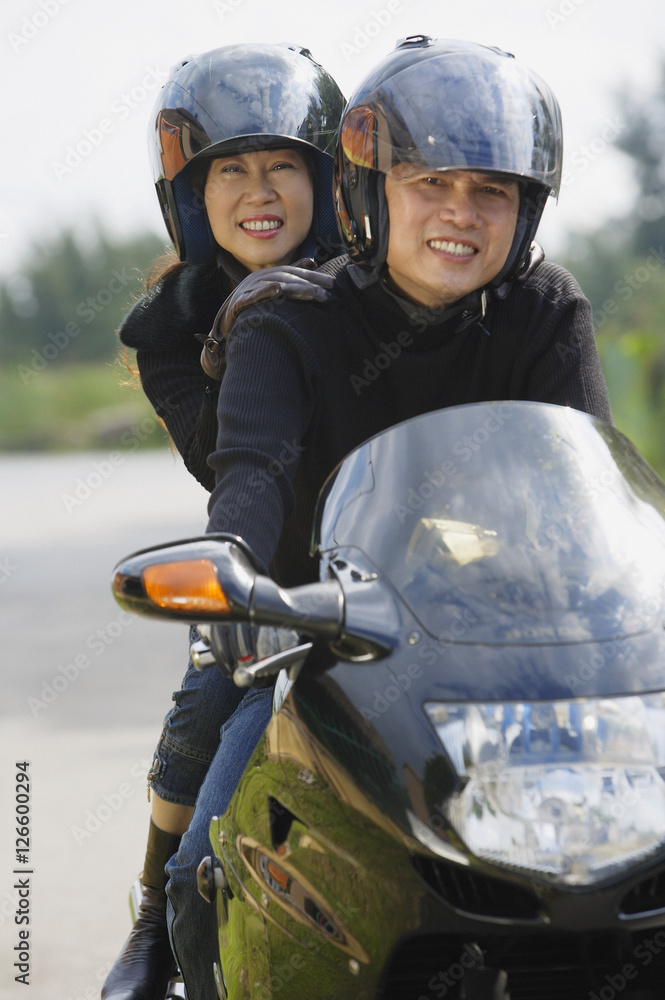 Man and woman riding motorcycle, wearing helmets, looking at camera ...