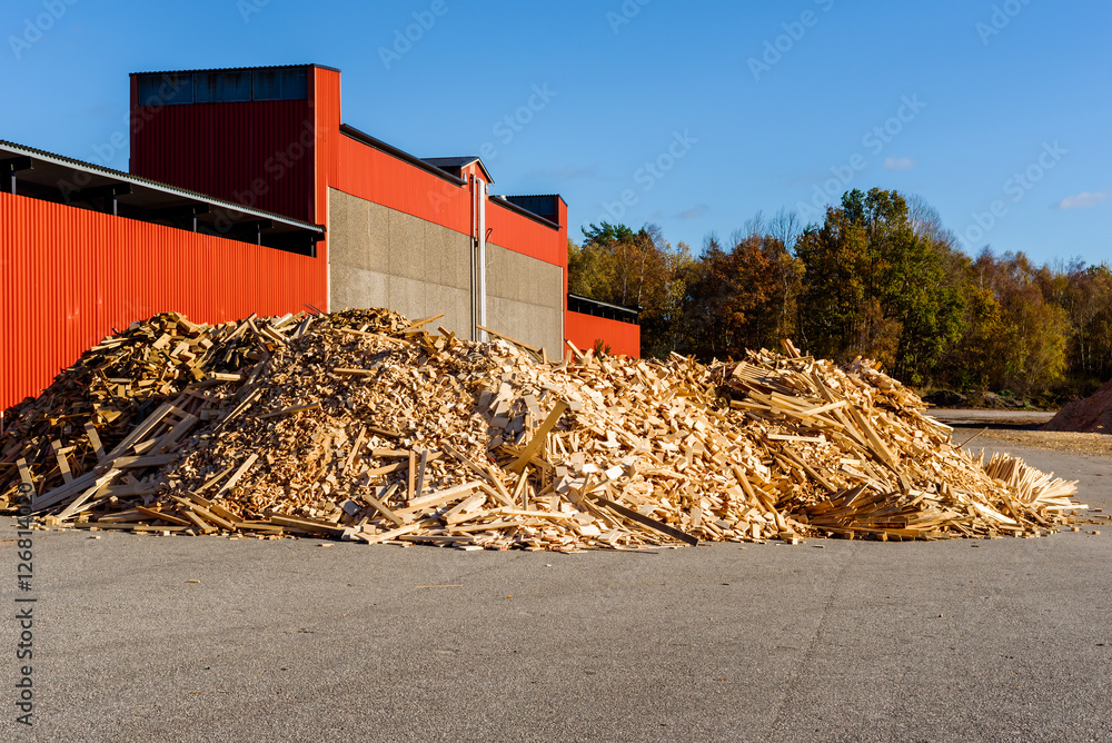 Pile of wooden debris in front of red industrial building. The wood is ...
