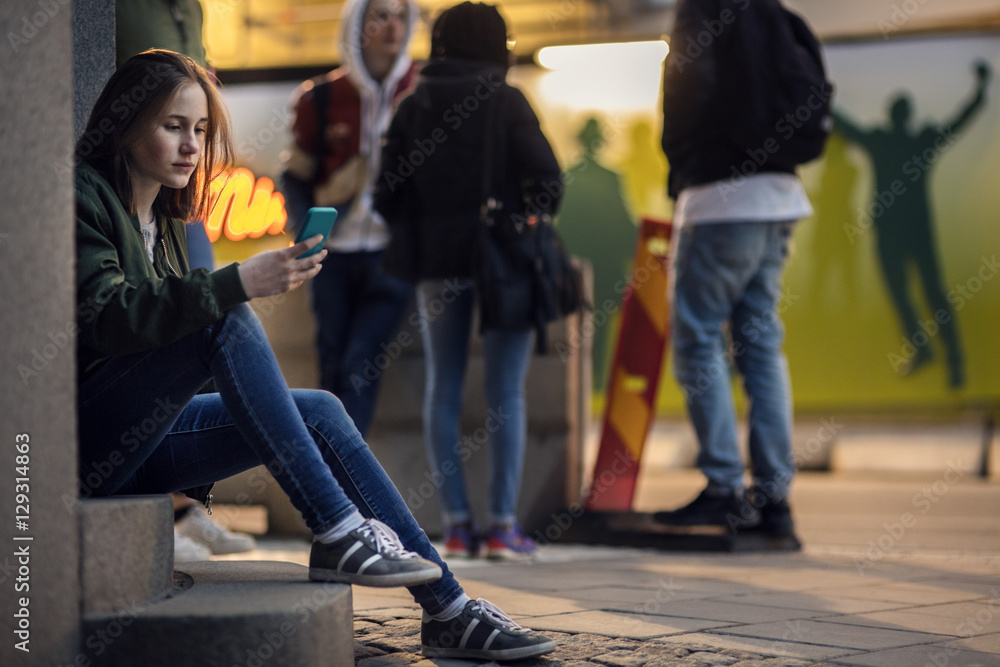 Thoughtful teenager using phone while sitting on steps against friends ...