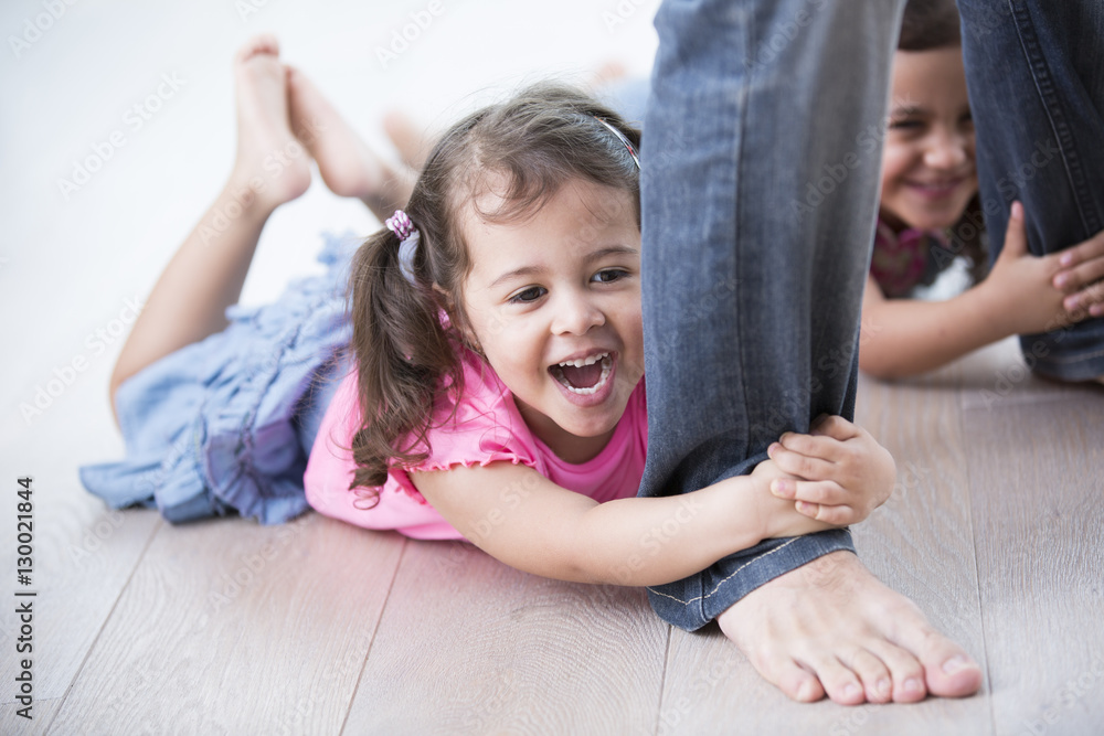 Playful girls holding father's legs on hardwood floor Stock Photo ...