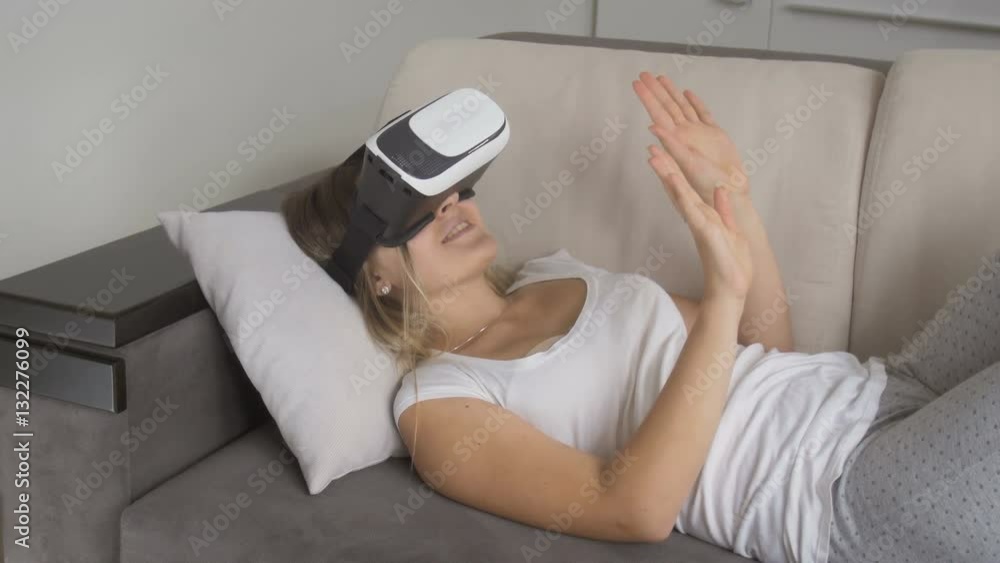 Young smiling woman lying on bed and watching 360 degree video in VR ...