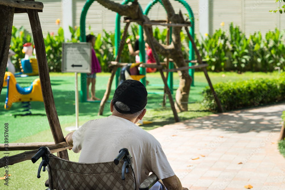 Old man sits on wheelchair looking at children playing on playground. Concept of youth recall ...