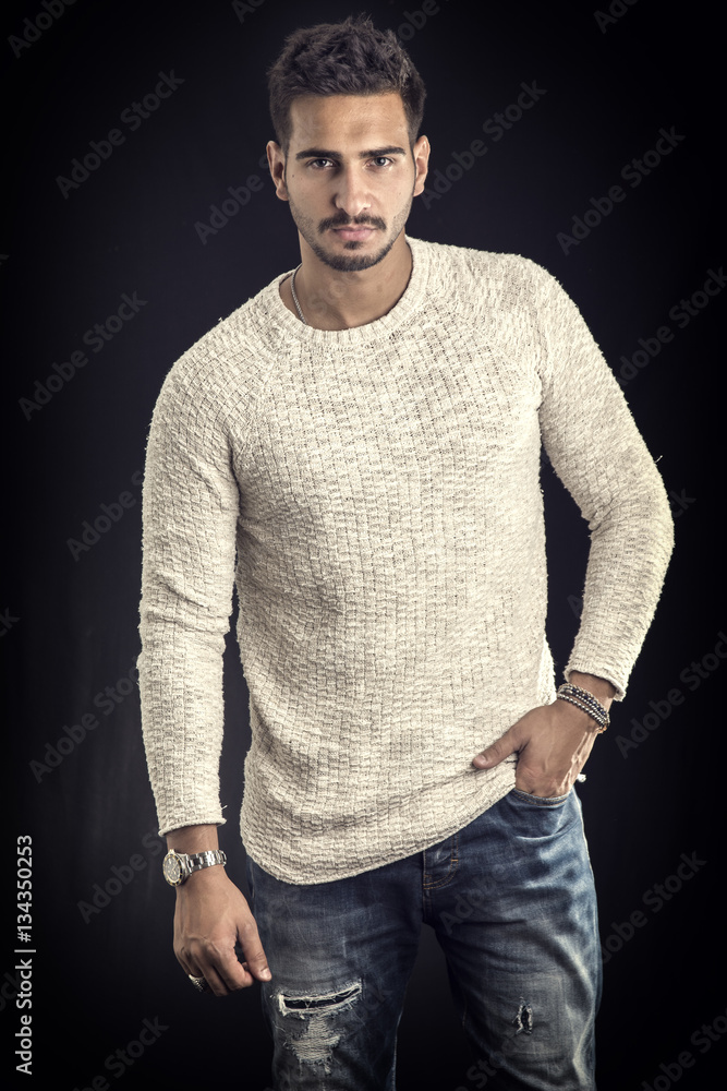 Handsome young man wearing white sweater and ripped jeans, on dark ...