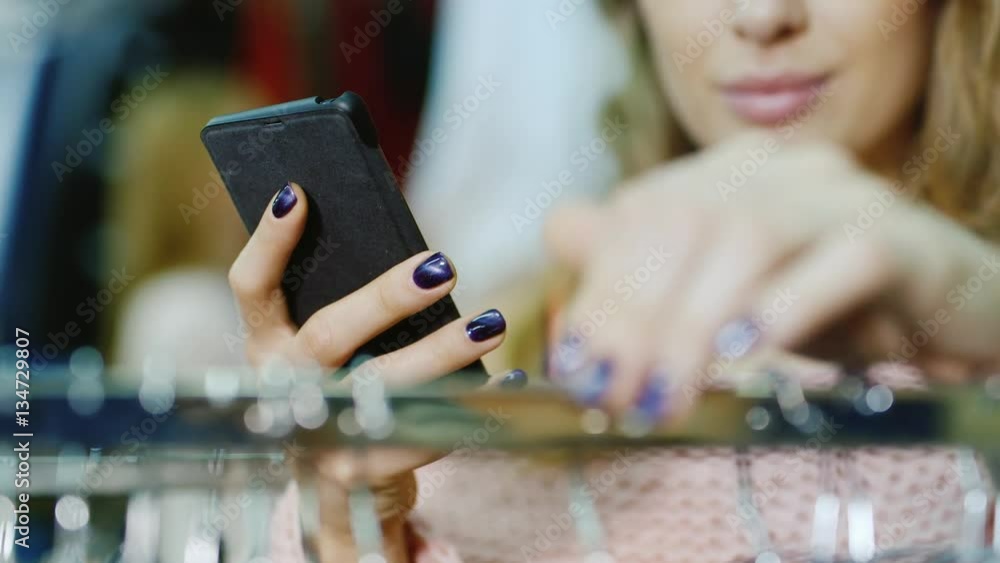 Woman chooses a dress in the store, looking at smartphone photo folder. In the frame of large ...
