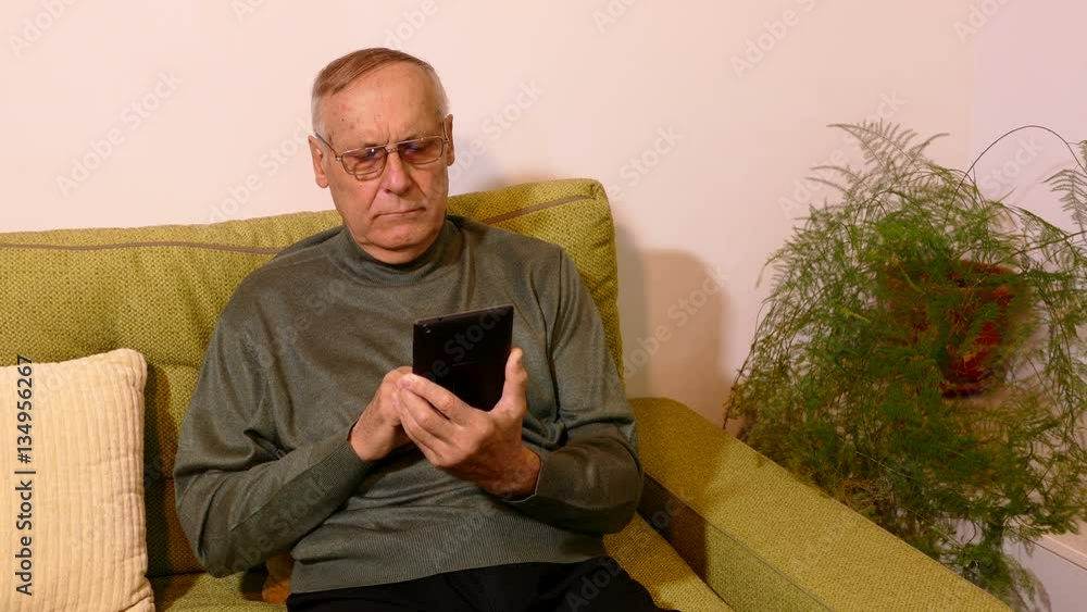 Old man sitting on the sofa in the living room and using his tablet ...