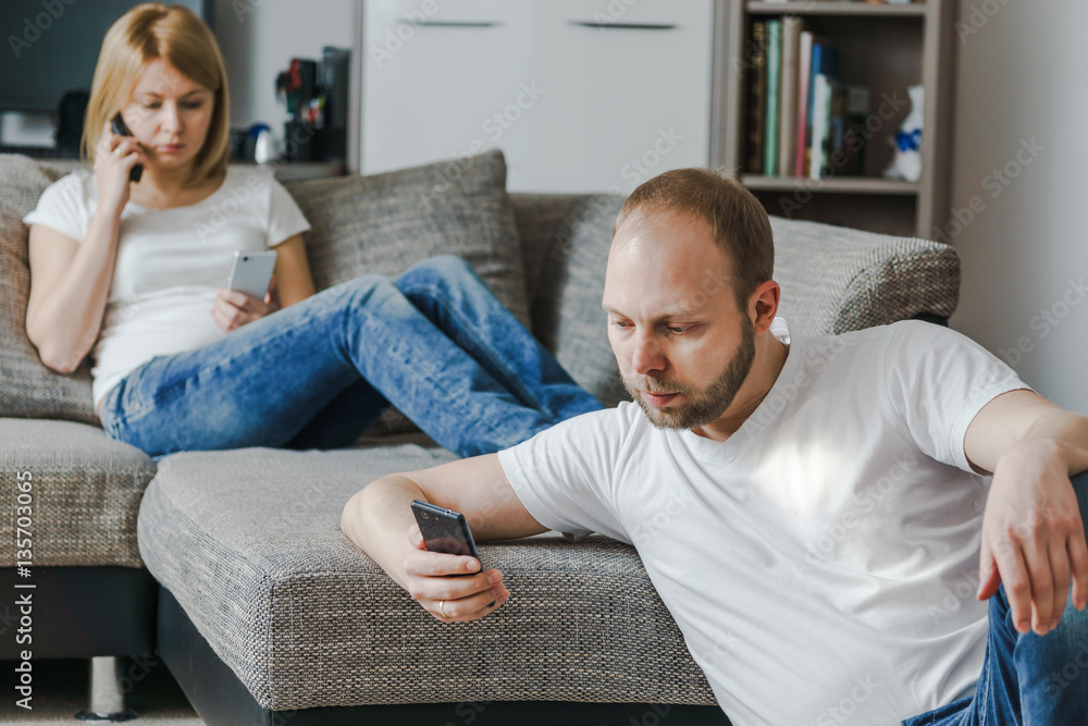 Young woman sitting at the sofa talking on phone while her husband is ...