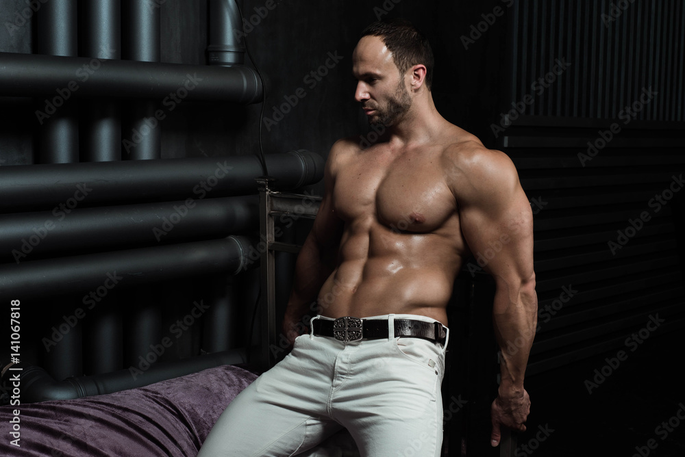 Sexy young shirtless handsome bodybuilder sits on a bed in a room with ...