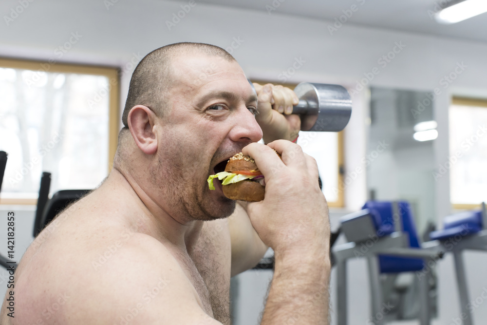 A big fat hungry man eats a hamburger with meat and cheese in the gym ...