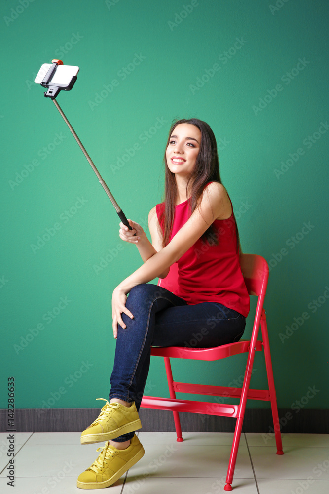 Beautiful young woman taking selfie while sitting on chair near color ...