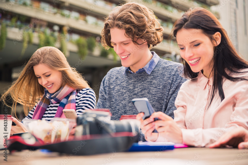 Smiling jolly students sit and look at their phone in time of a break ...
