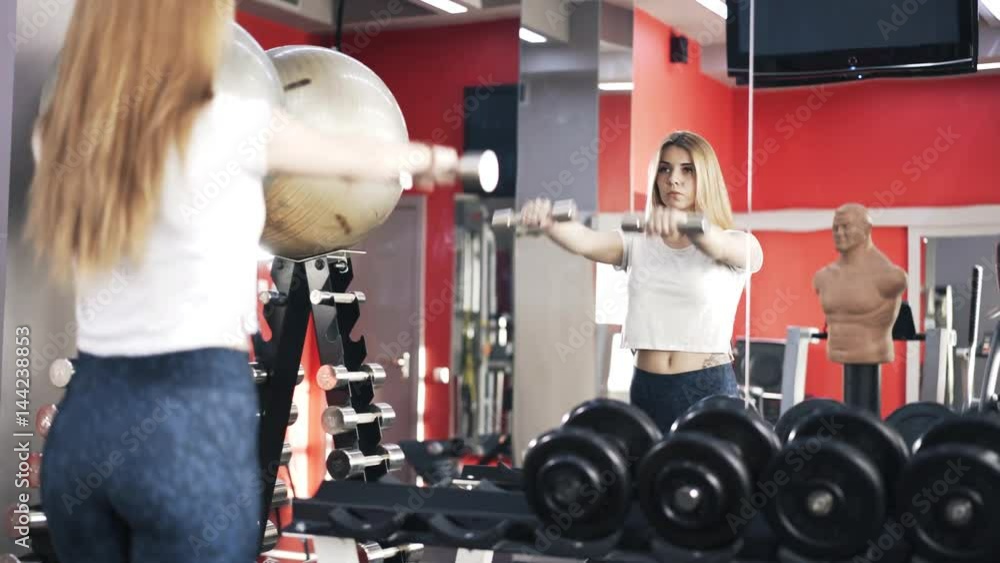 Reflection of a young blond woman wearing a white T-shirt doing a dumbbell exercise in a gym ...