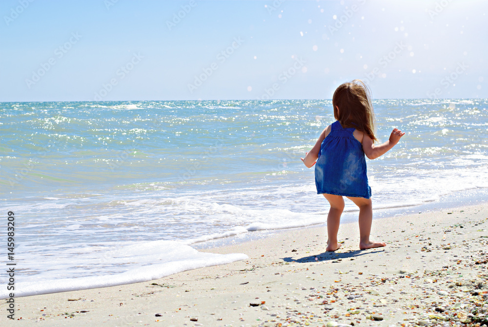 Cute little girl in denim dress shrowing stones or shells on the beach ...