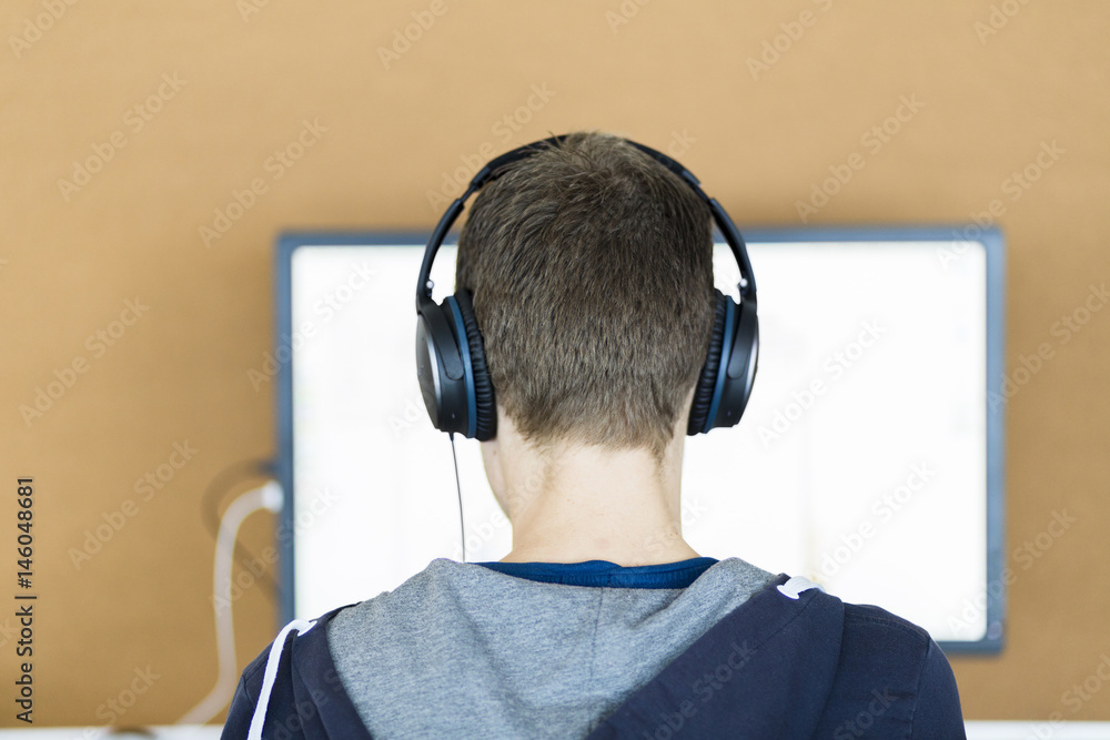 A young man behind a computer screen with headphones on, seen from the ...