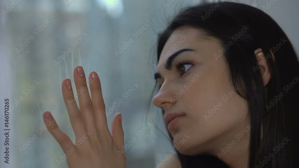 Brunette girl touches the glass with her hand and looks sad at the ...