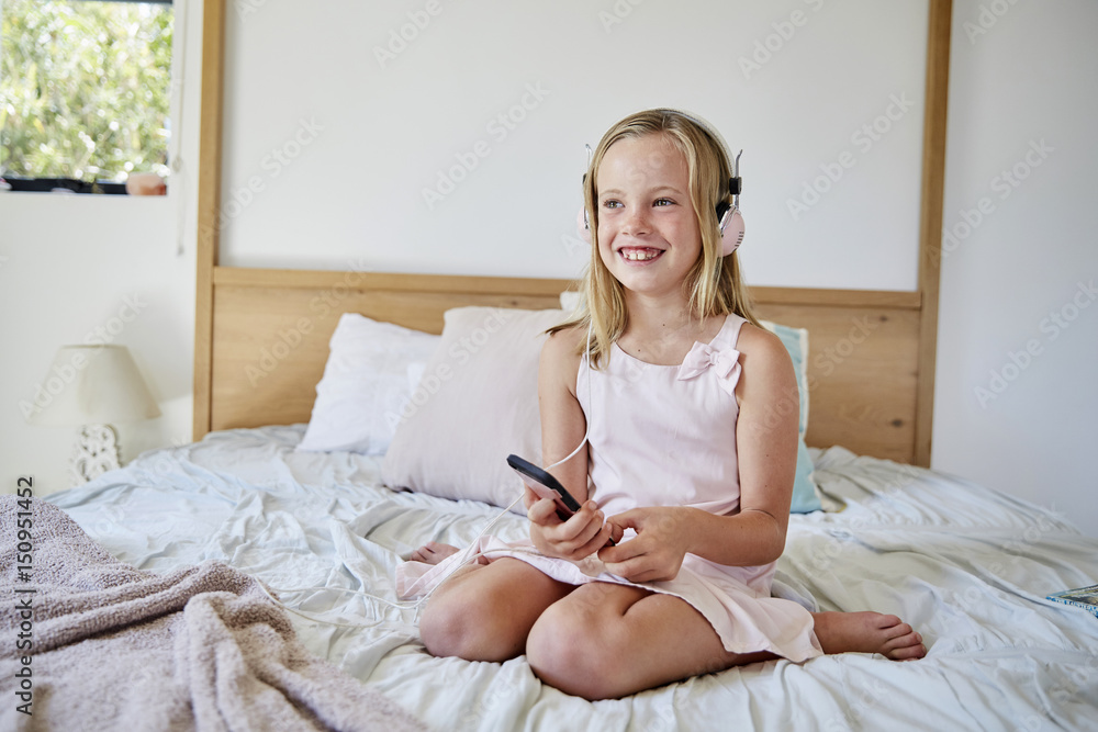 Portrait of smiling little girl sitting on bed listening music with ...