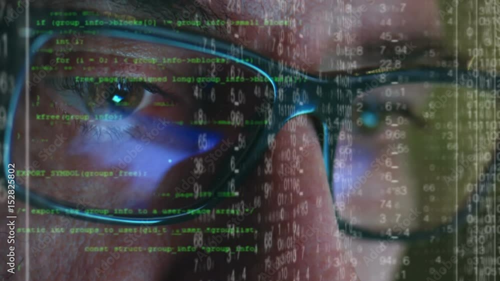 Male hacker working on a computer while green code characters reflect on his face in a dark ...