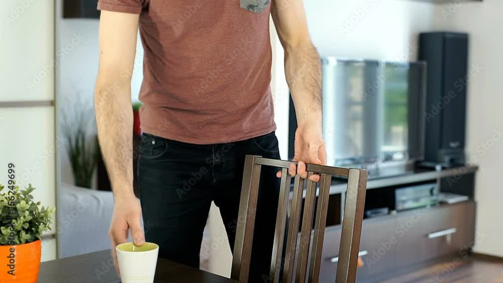 Irritated man sitting down by the table in the flat and drinking coffee ...