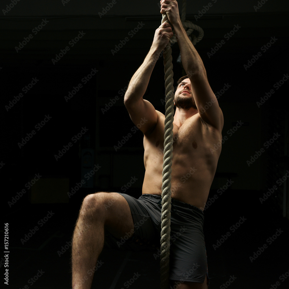 Male athlete or sportsman climbing rope at dark gym. Topless handsome ...
