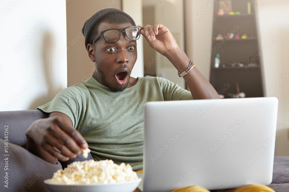 Horrified Afro American man dressed casually sitting at home watching ...