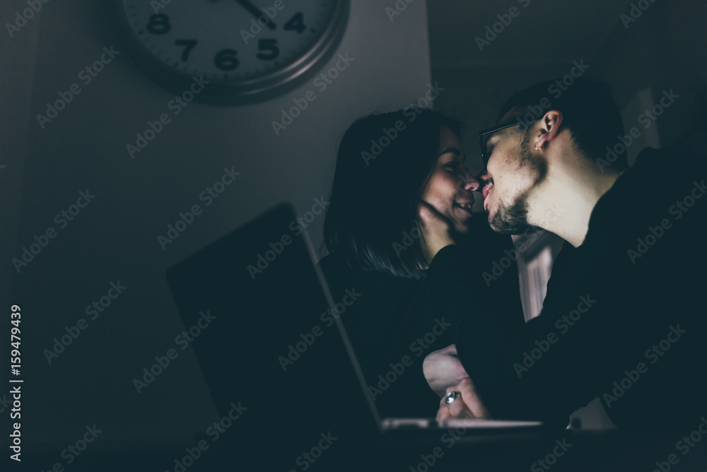 Young couple kissing using computer illuminated by screen light smiling ...