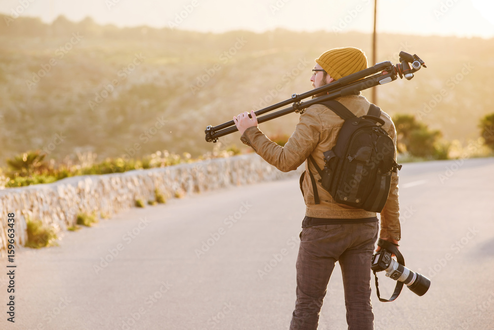 Young photographer walking on desert road with tripod on his shoulder ...